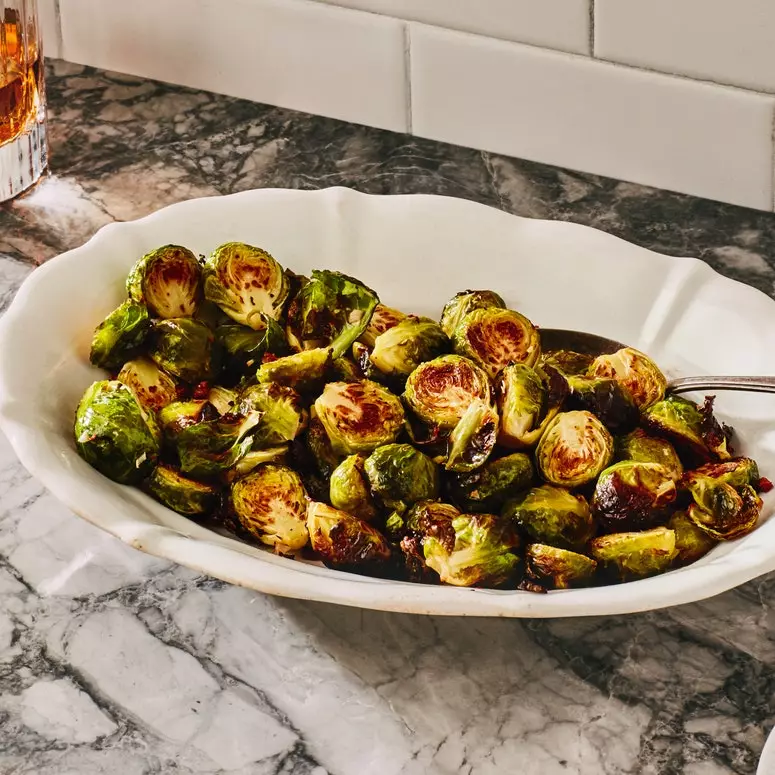 Ti yan Brussels Sprouts pẹlu ata ilẹ ati Pancetta ninu satelaiti pẹlu ṣibi ti o n ṣiṣẹ ati awọn ohun mimu ni ẹgbẹ