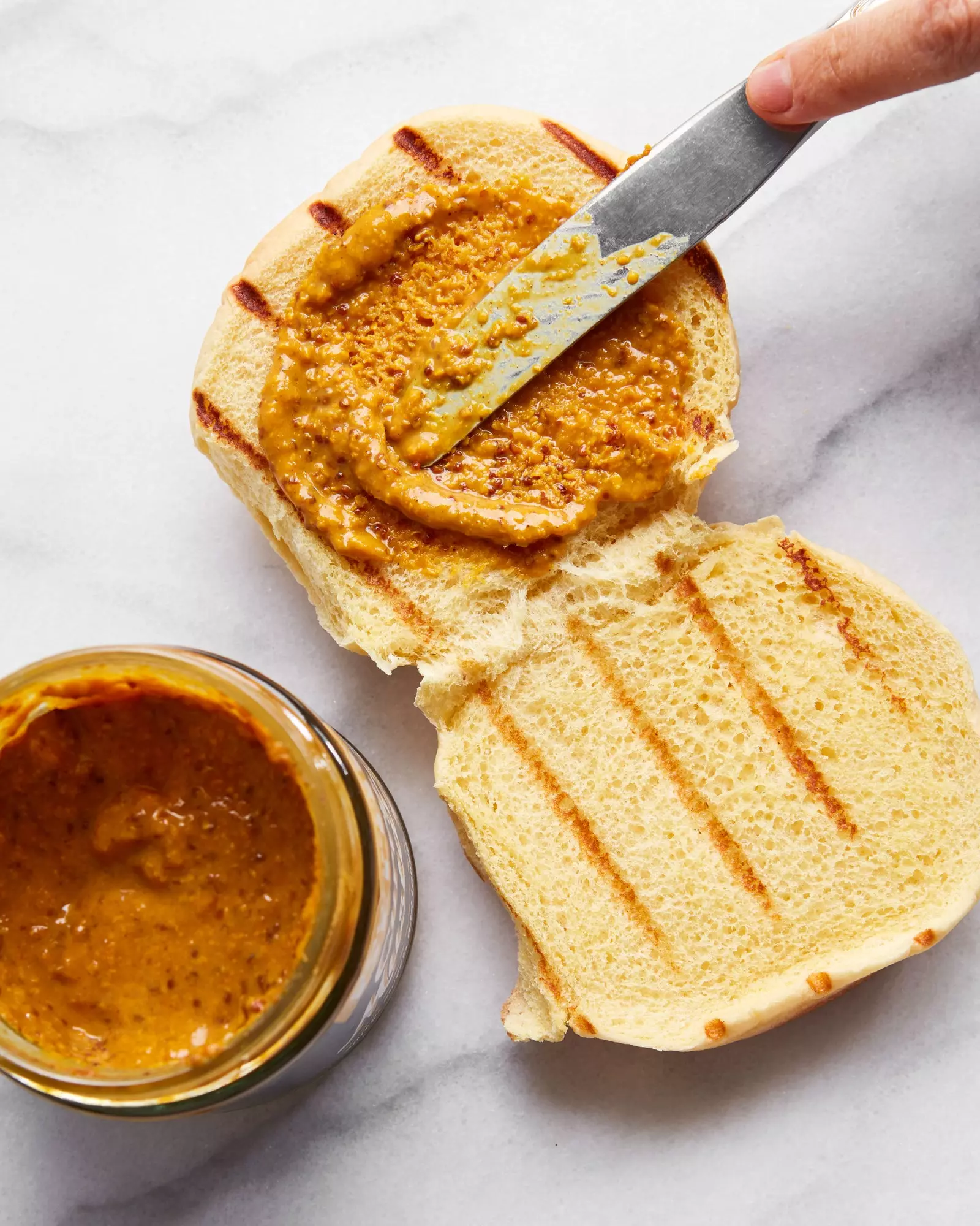 Foto seseorang sedang menebar sawi kari pada roti burger panggang.