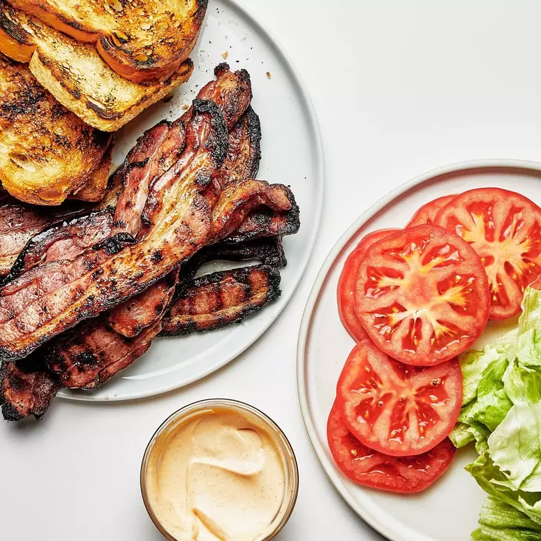 Prvek BLT slaniny toastový chléb nakrájená rajčata natrhaný salát a majonéza vše rozložené na dvou talířích.