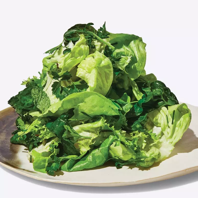 En haug med blandede grønnsaker og urter stablet høyt på en salattallerken i keramisk jordtone på hvit bakgrunn.