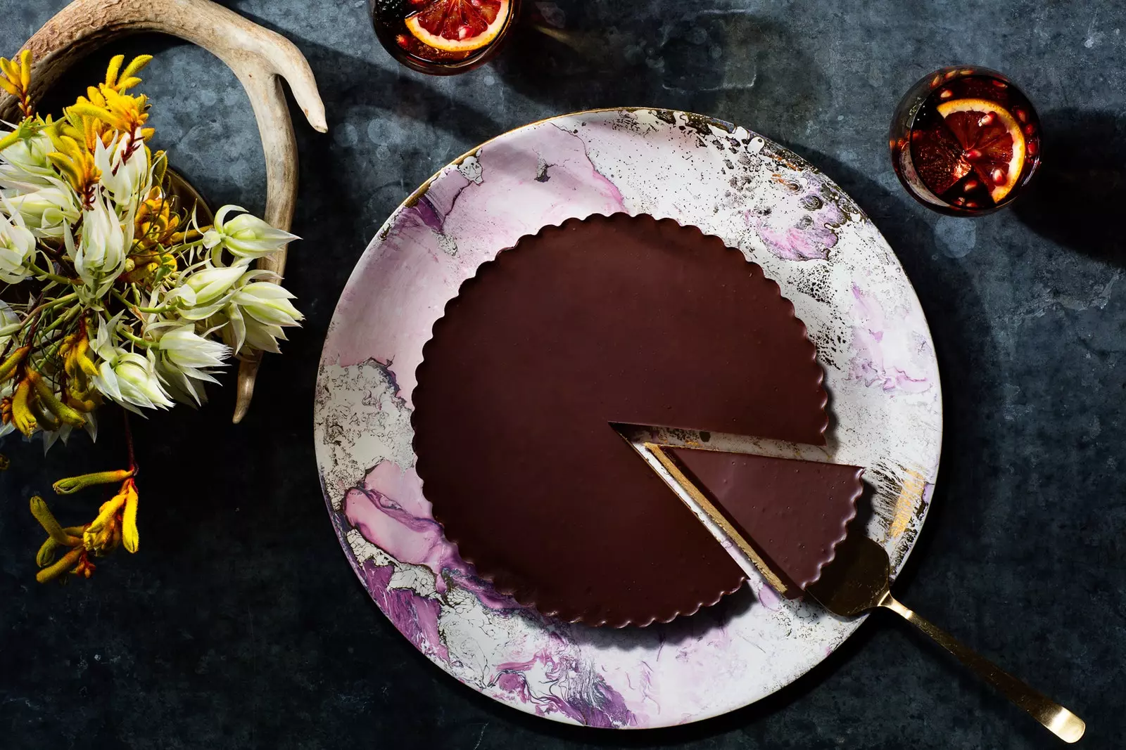 Óriás csokoládé mogyoróvajas csésze rózsaszín tányéron, két pohár ital és virágok sötétszürke háttér előtt.