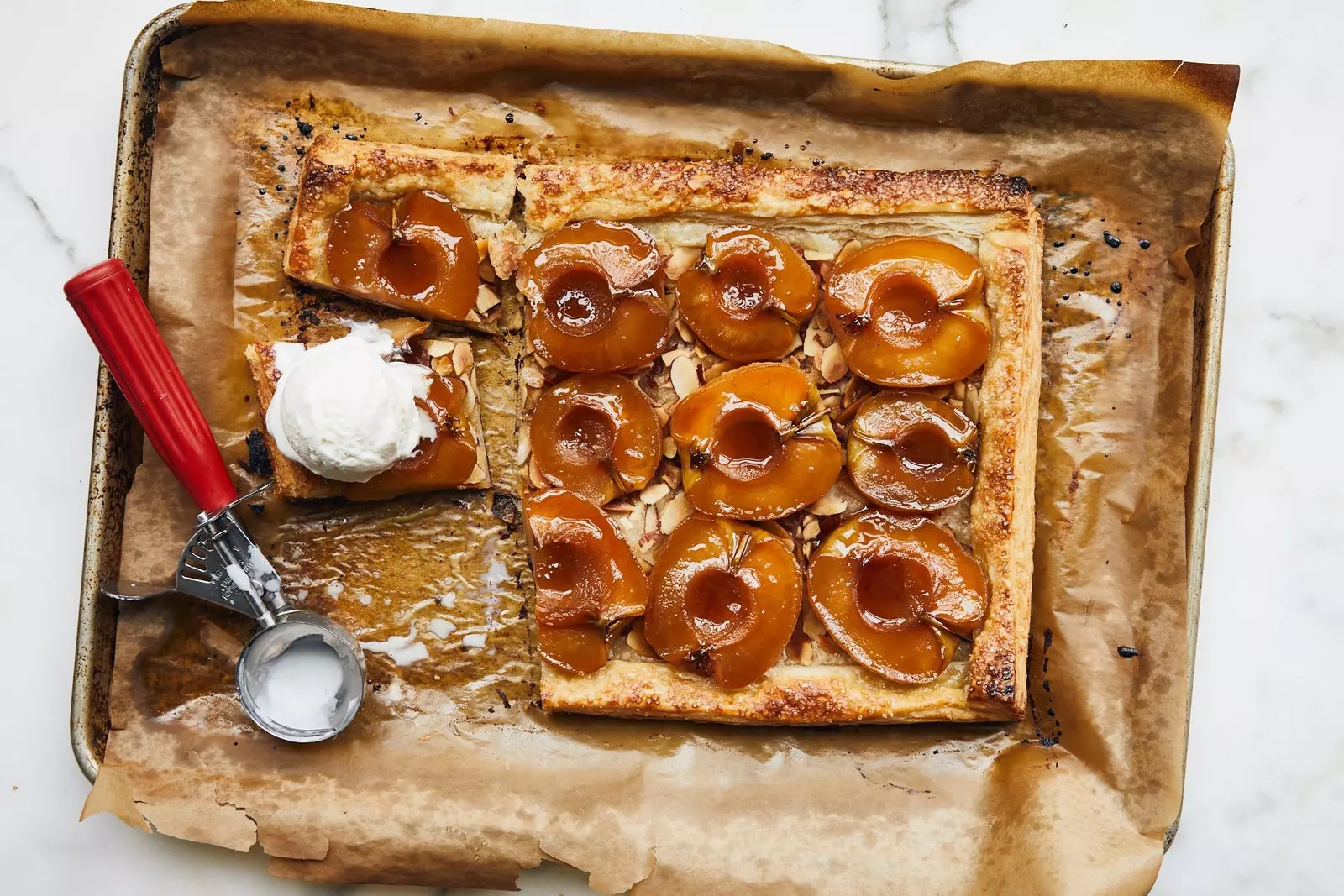 Prostokątna tarta z ciasta francuskiego zwieńczona połówkami głęboko karmelizowanych jabłek i pokrojonymi migdałami. Lewa trzecia tarta...