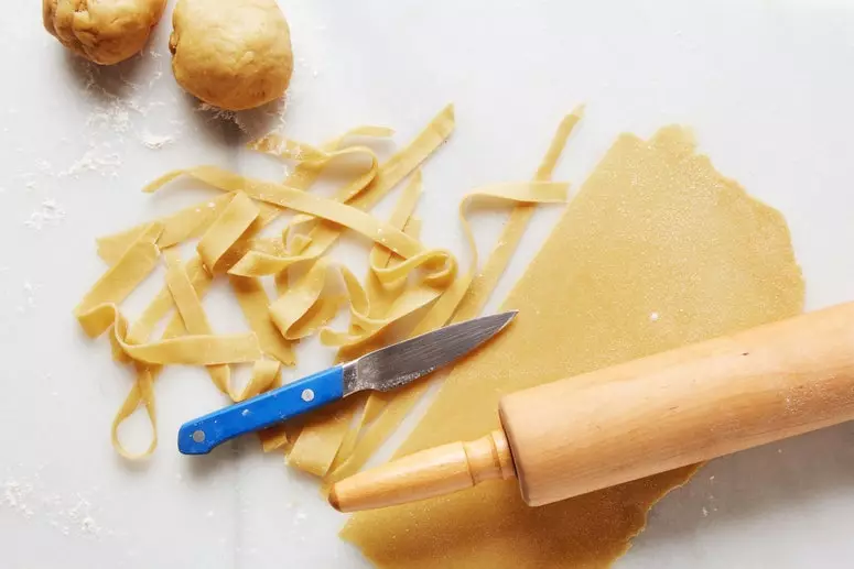 Foto de masa de pasta cortada en tiras finas con un cuchillo de cocina, un rodillo de madera y dos bolas de masa a un lado.